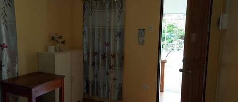 Um quarto pequeno com uma mesa de madeira, um armário, uma cortina florida e uma porta que dá para o exterior.