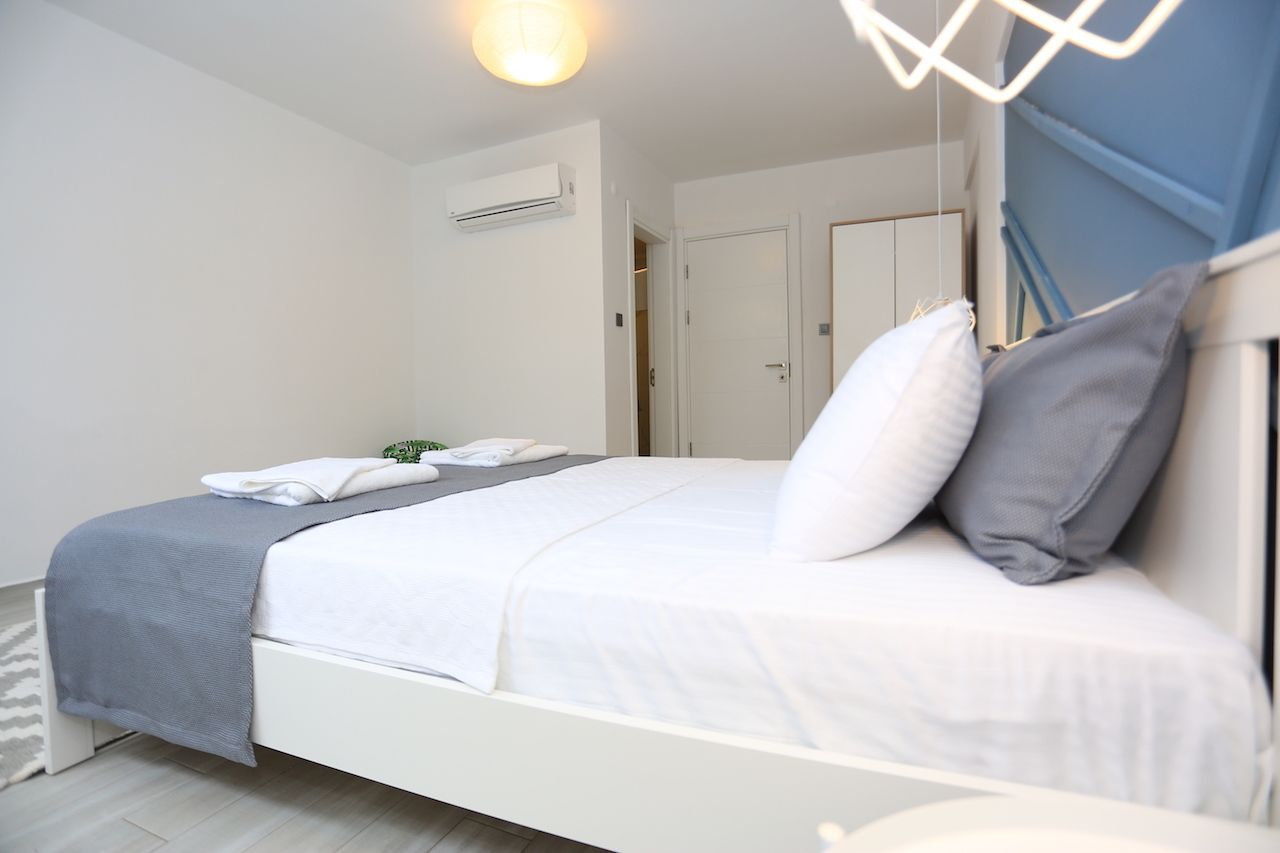 Quarto moderno com cama branca, almofadas cinzas e brancas, manta cinza e mesa de cabeceira branca.