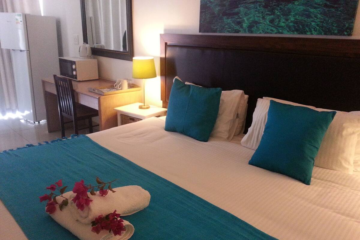 Quarto de hotel com cama, escrivaninha, cadeira, abajur e um quadro na parede.