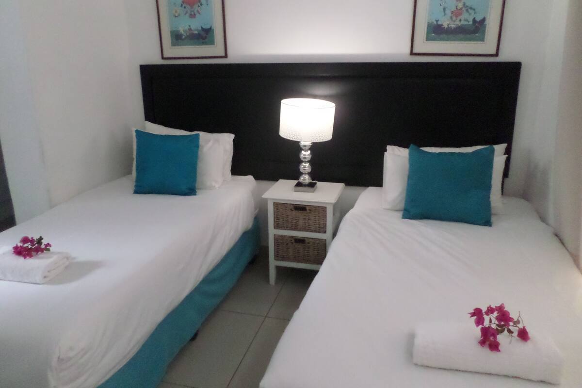 Quarto de hotel com duas camas de solteiro, cada uma com um travesseiro azul e edredom branco. Há uma mesa de cabeceira com uma luminária entre as camas.