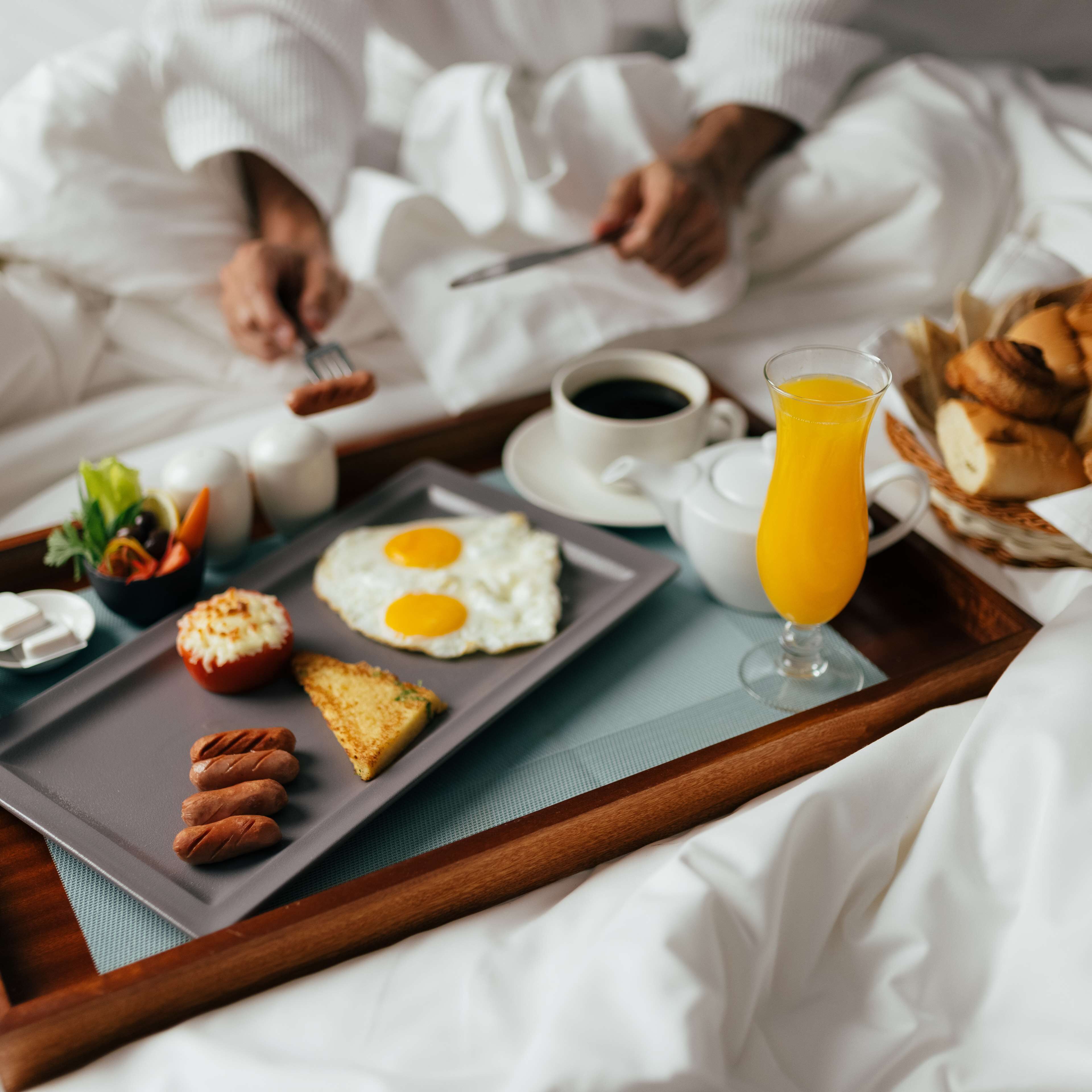 Bandeja de café da manhã com ovos, torradas, linguiça e um copo de suco de laranja sobre a cama.