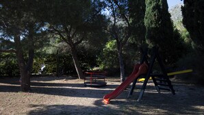 Um parquinho infantil com um escorrega vermelho e estruturas de escalada, cercado por árvores e um piso de cascalho.