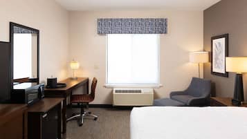 Quarto de hotel com uma cama, uma escrivaninha com um computador, uma cadeira, um abajur e uma janela com cortina decorada.