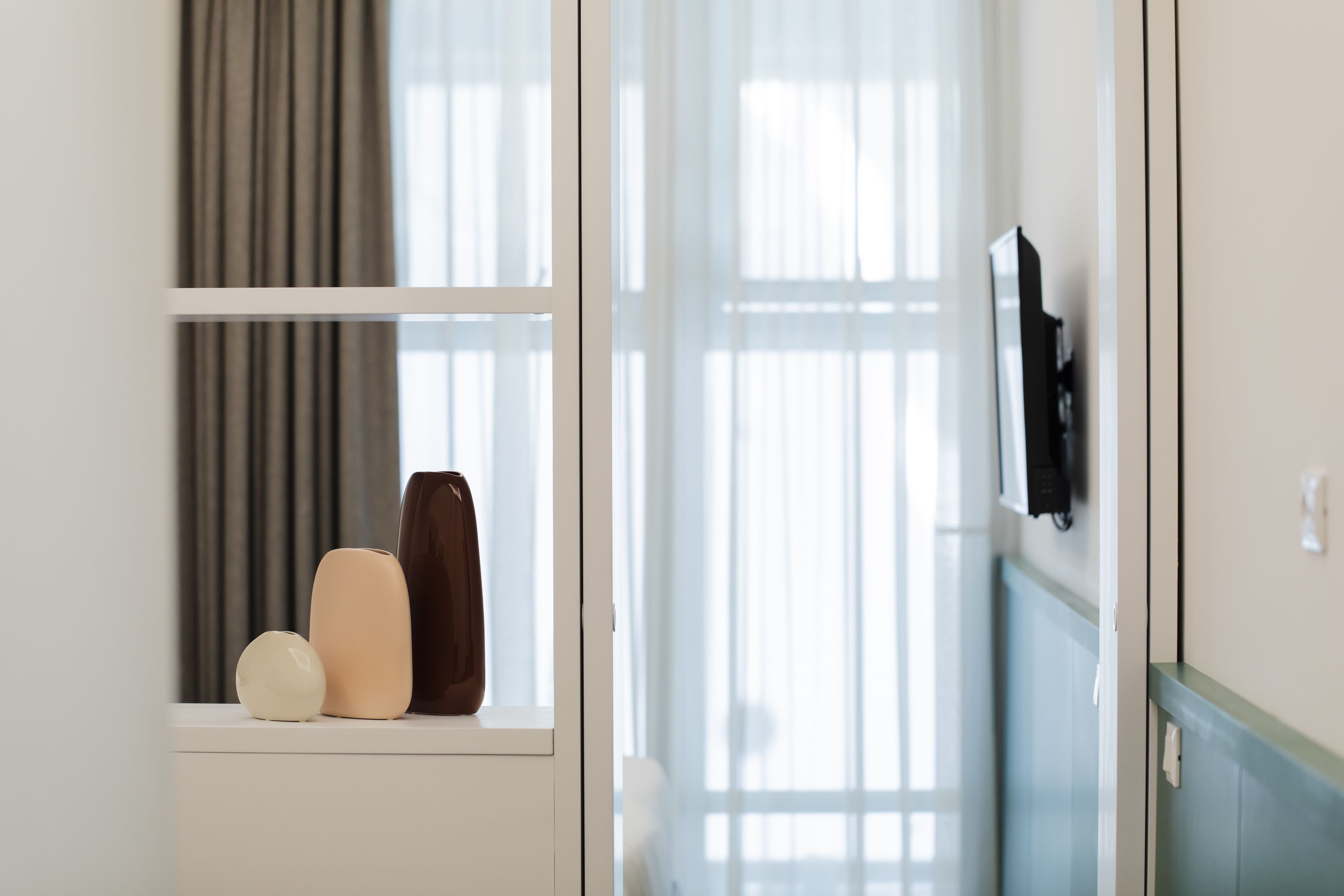 Quarto de hotel com janela, TV de tela plana na parede e uma pequena mesa com três vasos decorativos.