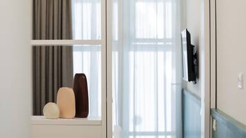 Quarto de hotel com janela, TV de tela plana na parede e uma pequena mesa com três vasos decorativos.