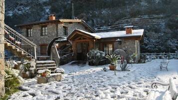 Uma casa de pedra com uma escadaria de madeira que leva à entrada, rodeada por chão coberto de neve e plantas.