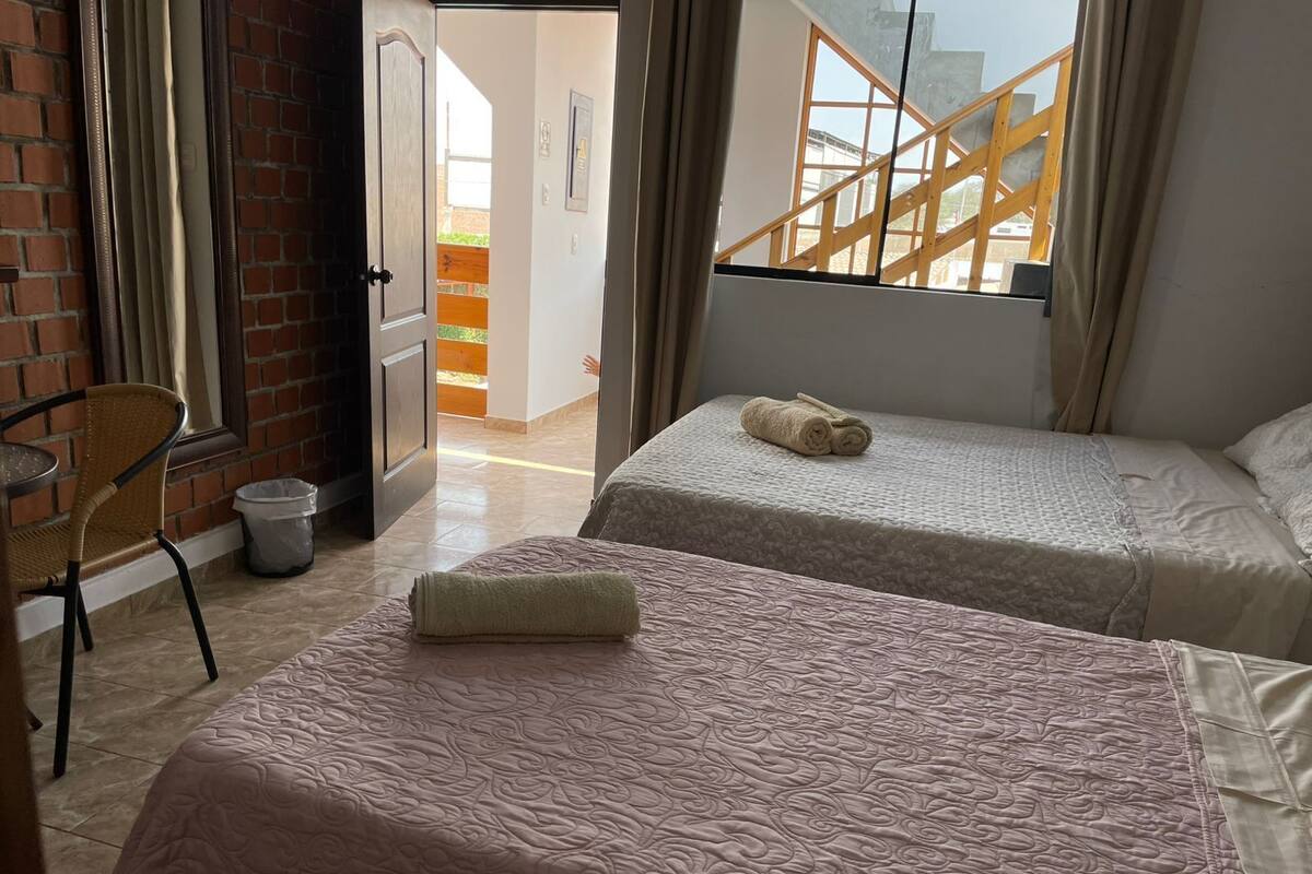 Quarto de hotel com duas camas, uma cadeira, um cesto de lixo e vista para uma escada de madeira lá fora.