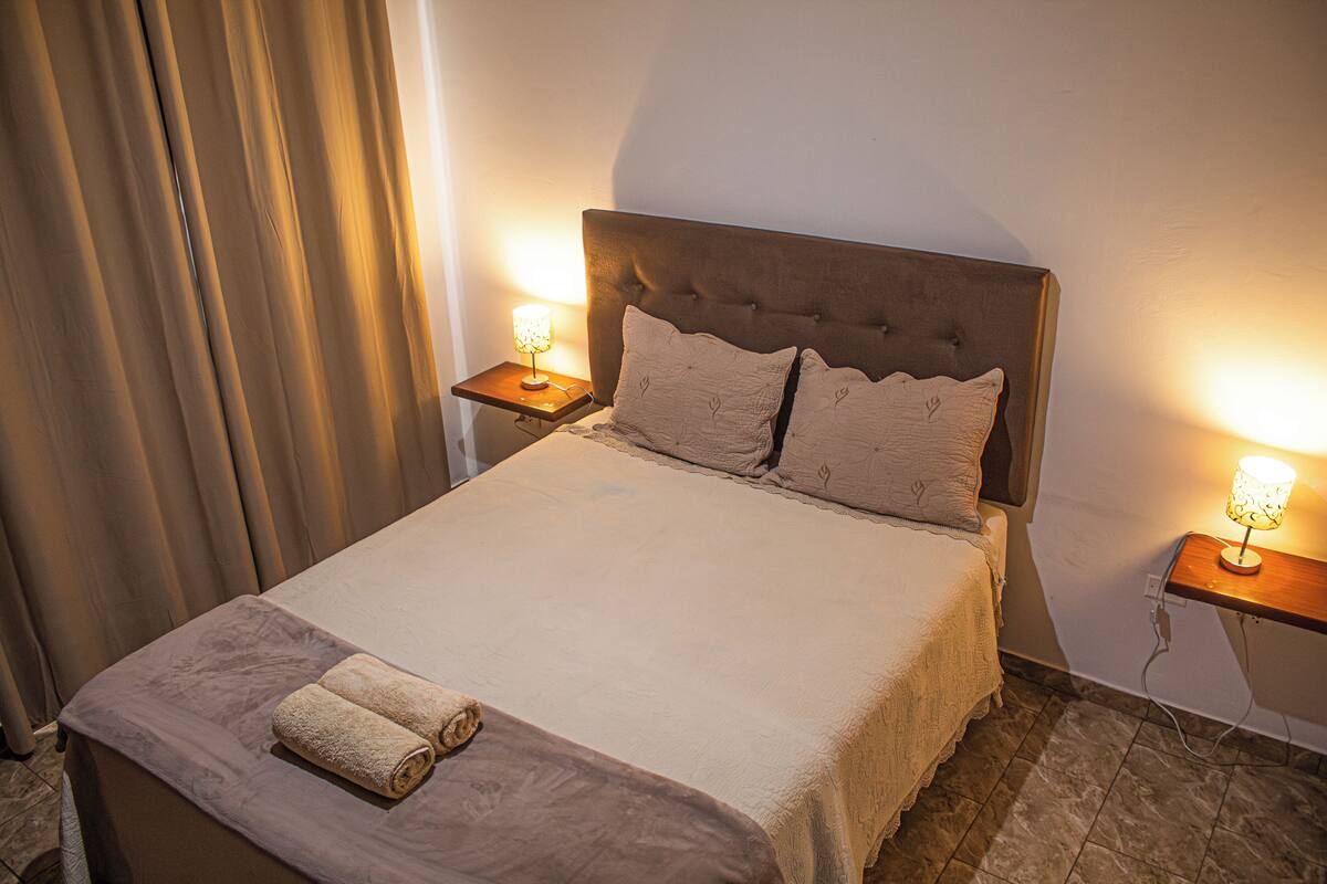 Cama bem arrumada com cabeceira, duas luminárias de apoio e uma toalha enrolada sobre a cama.