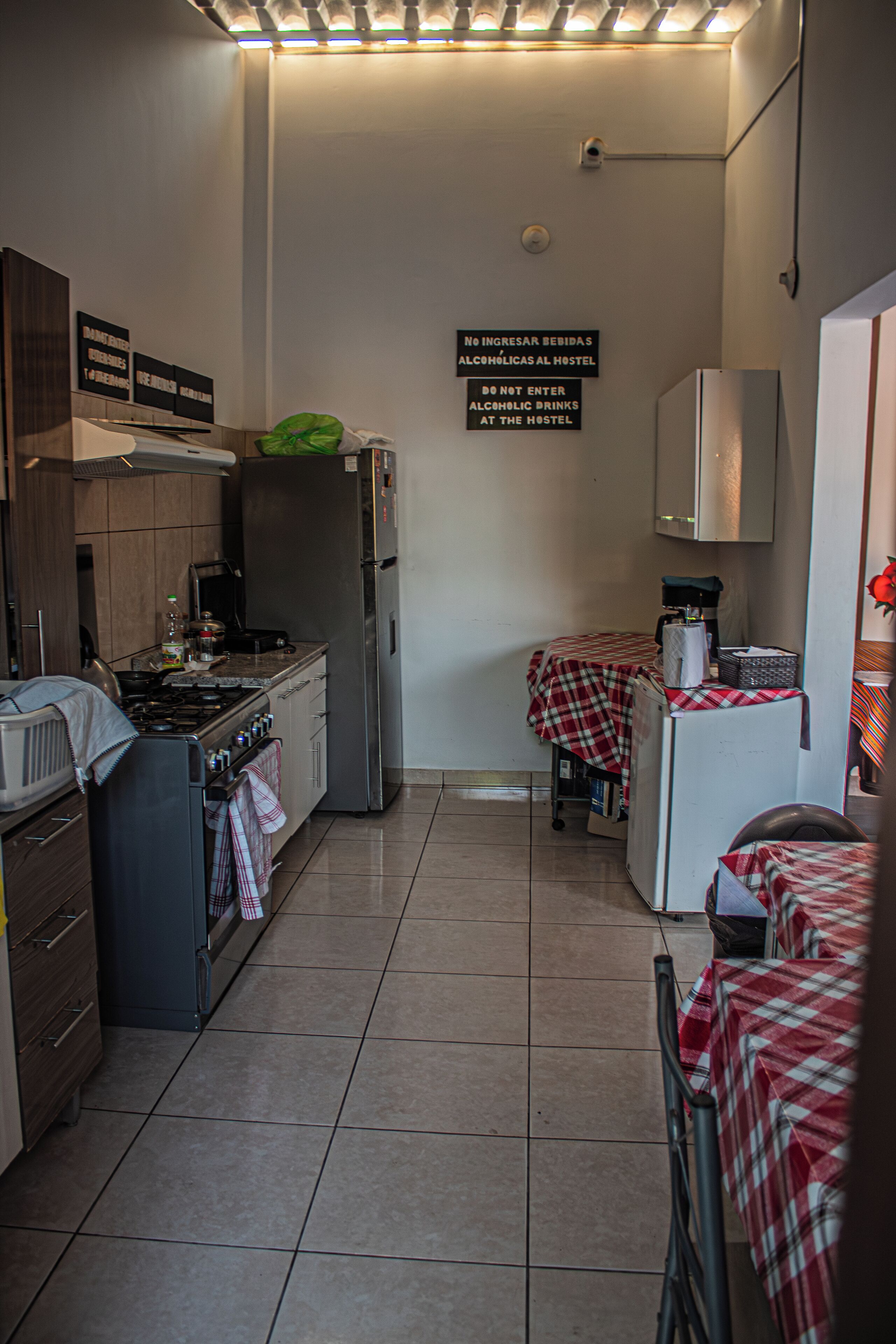 Uma cozinha compacta com fogão, geladeira e uma mesa com toalha xadrez. Um aviso na parede proíbe bebidas alcoólicas.