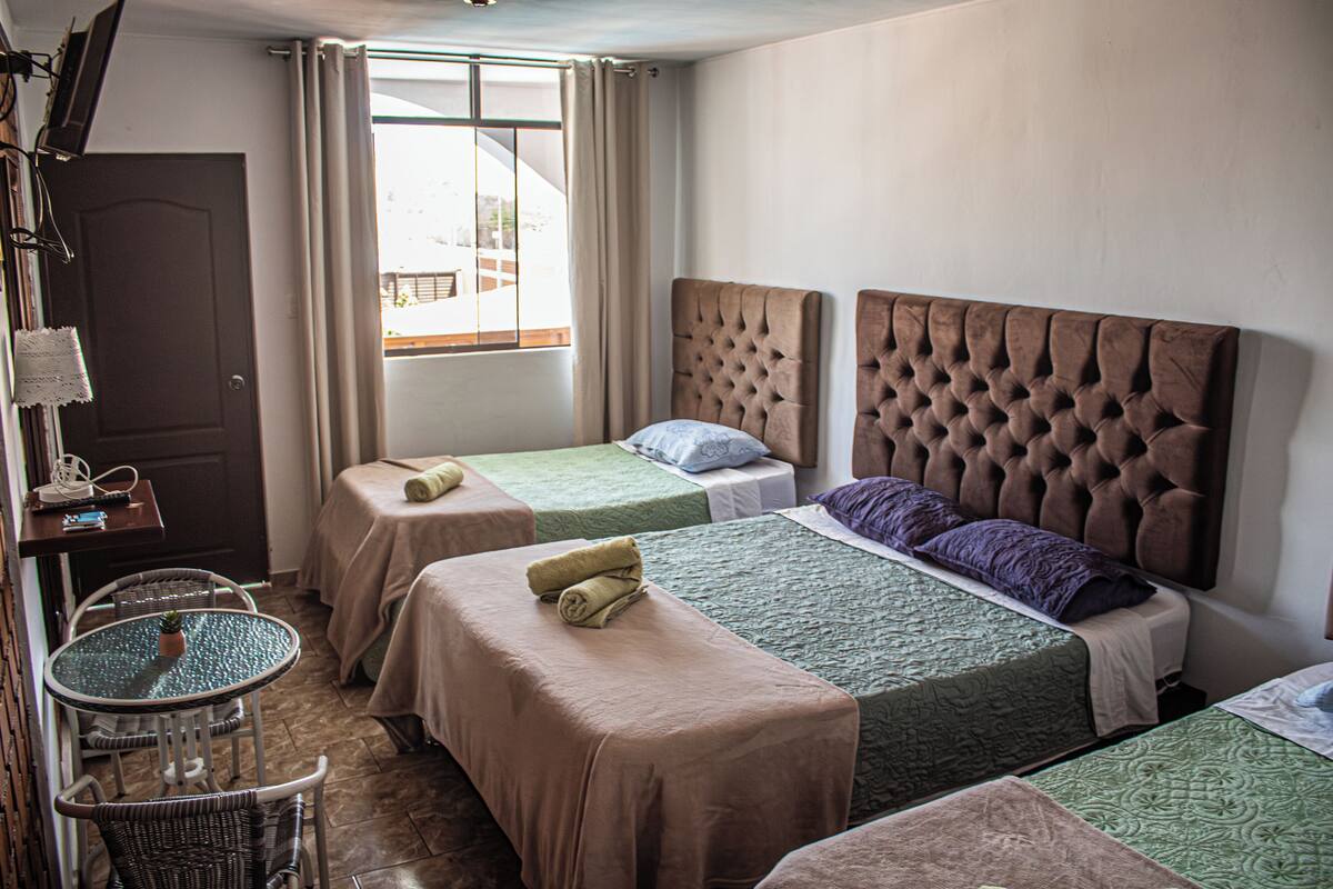 Quarto de hotel com duas camas de solteiro, uma pequena mesa com cadeiras, uma escrivaninha com abajur e uma janela com cortinas.