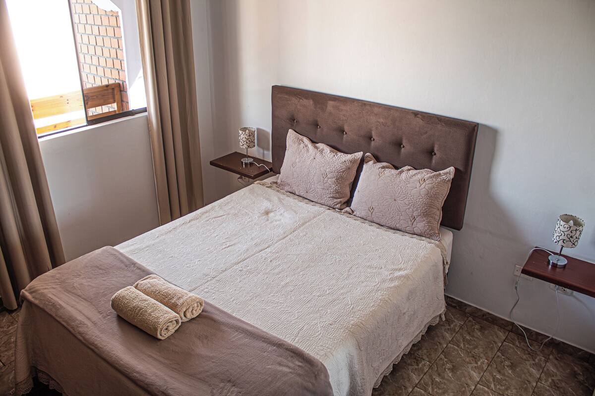 Uma cama bem arrumada com cabeceira marrom, uma mesa de cabeceira com luminária e uma janela com cortinas.
