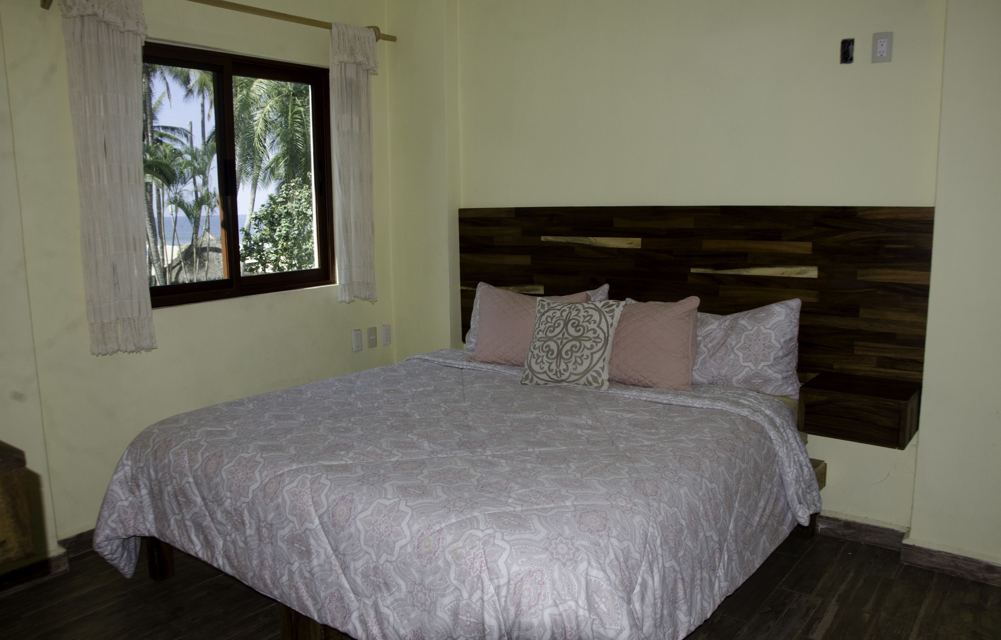 Quarto com cama grande, janela com cortinas e vista para palmeiras.