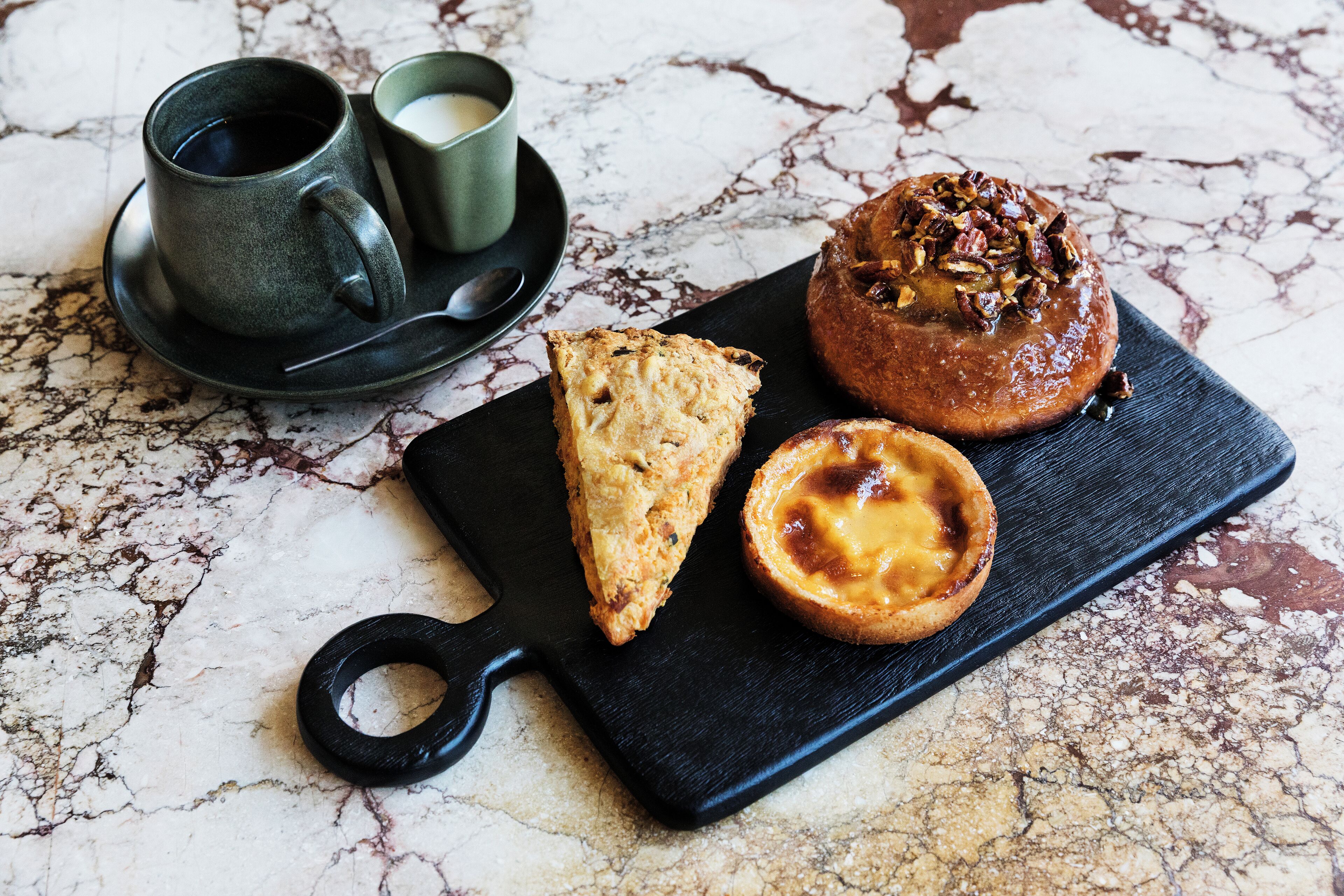 Uma tábua de servir preta com um doce, uma fatia de pão e uma xícara de café sobre uma superfície de mármore.