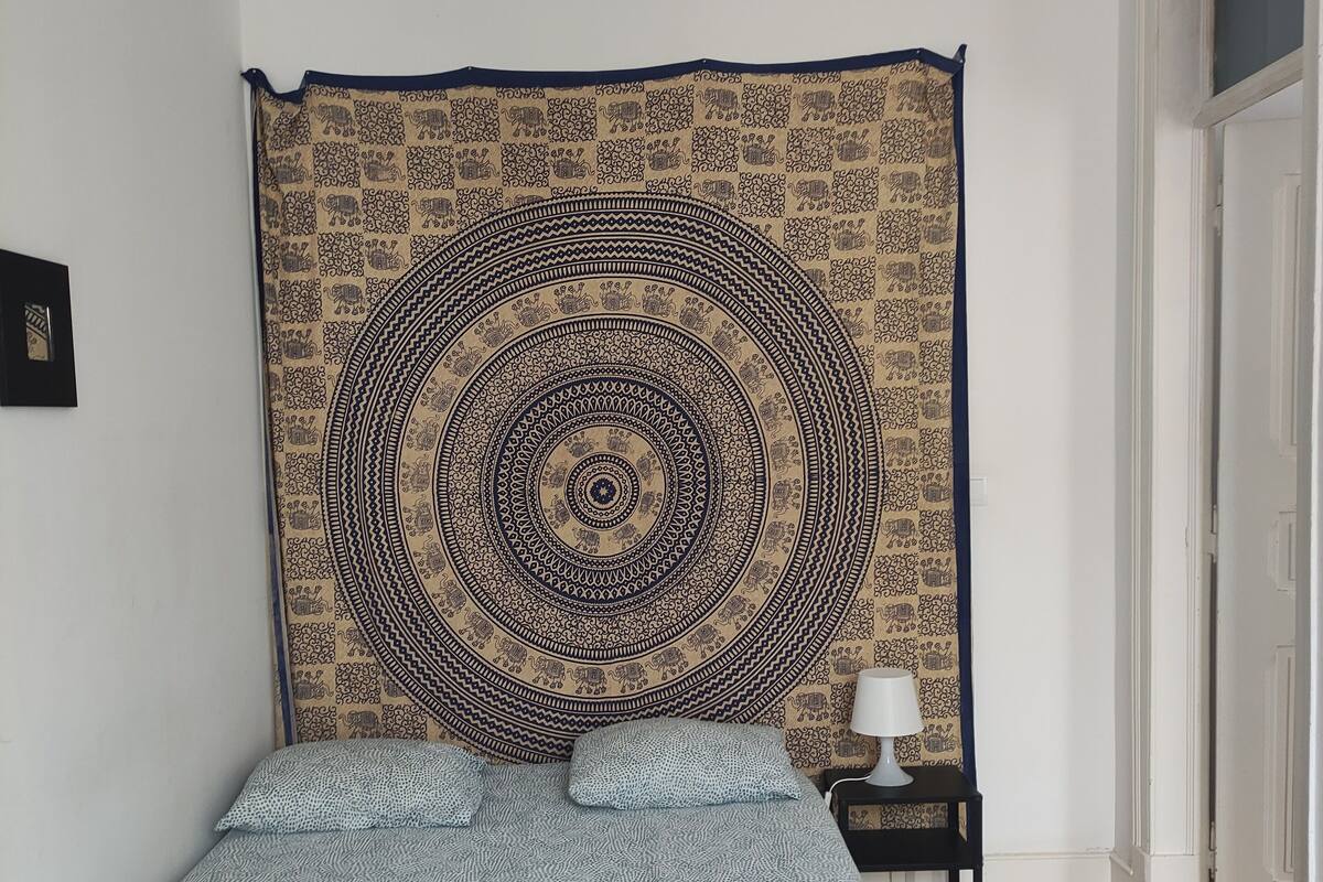Quarto com tapeçaria estampada, cama com roupa de cama estampada, toalha enrolada, mesa de cabeceira com abajur e piso de madeira.