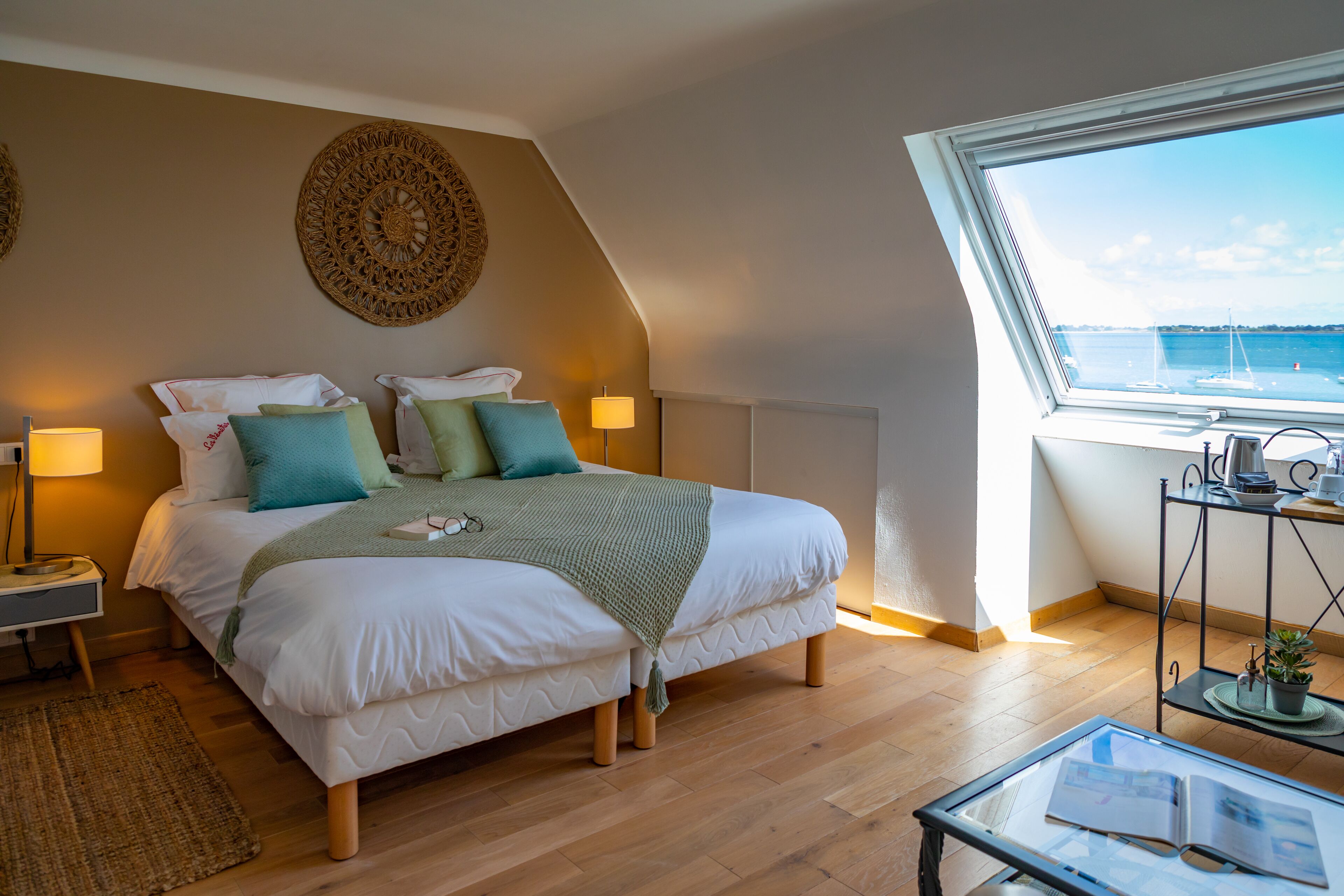Quarto com uma cama grande, vista para o mar pela janela, mesa de cabeceira com abajur e uma mesinha com um livro.