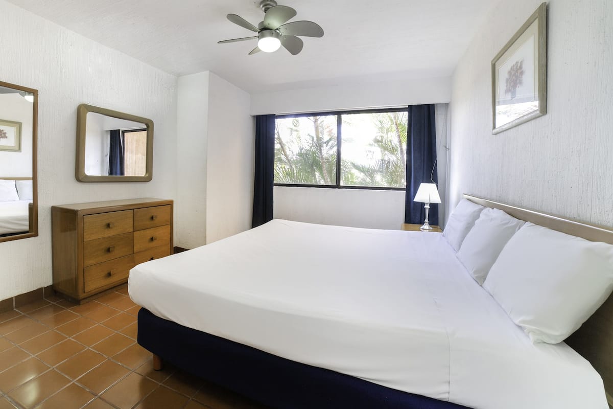 Quarto de hotel com uma cama, cômoda, ventilador de teto, janela com cortinas e um quadro na parede.