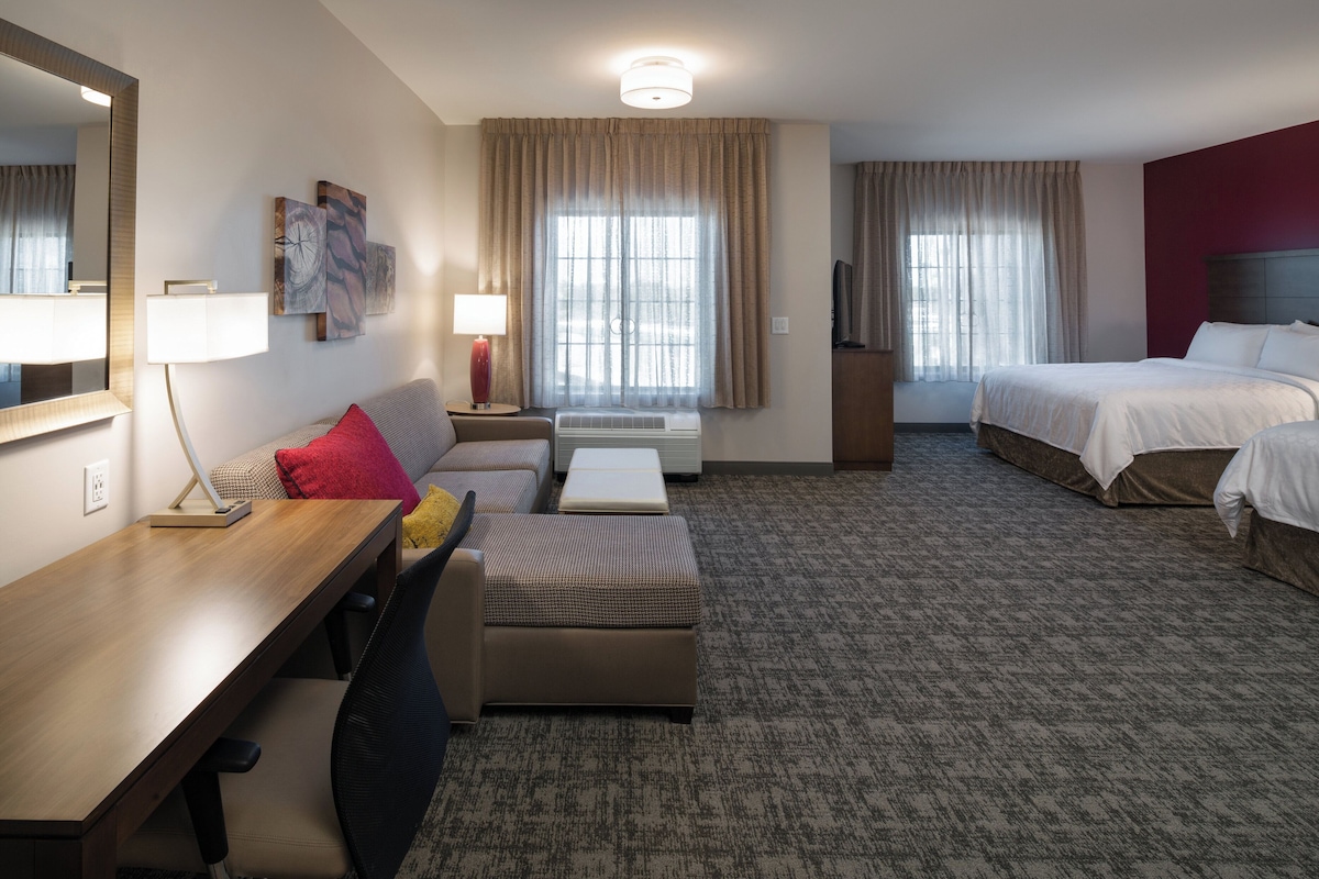 Quarto de hotel com cama, escrivaninha, cadeira, sofá, televisão e janela com cortinas.