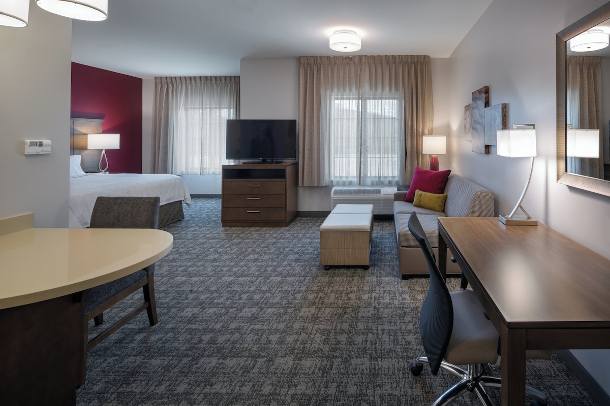 Quarto de hotel com cama, escrivaninha, cadeira, televisão, luminária e janela com cortinas.