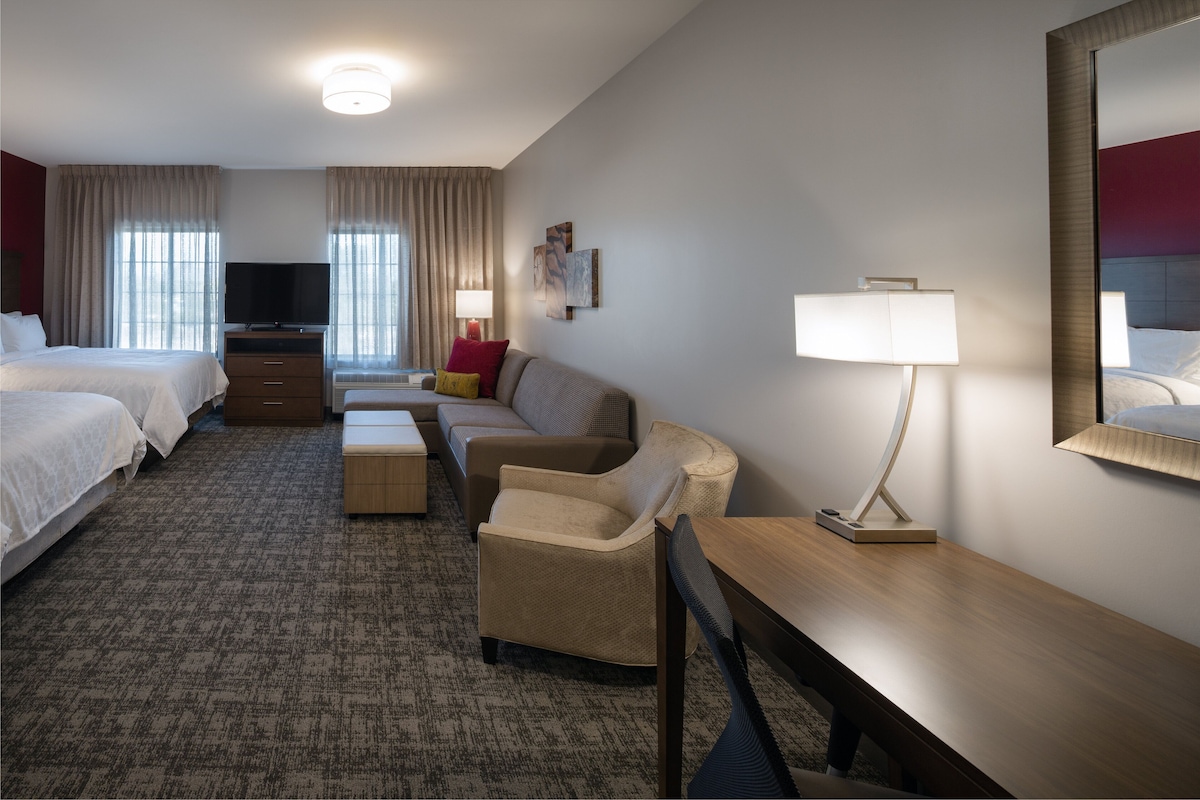 Quarto de hotel com uma cama, sofá, escrivaninha, televisão e luminária.