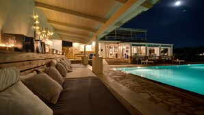 Uma área à beira da piscina com deck de madeira, piscina e um espaço de estar coberto com almofadas.