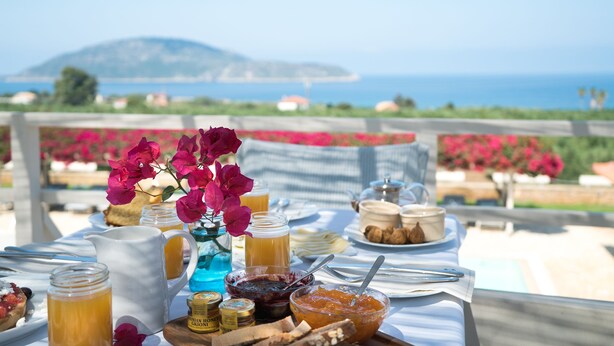 Uma mesa posta com itens de café da manhã, incluindo potes de geleia, um jarro e um vaso com flores, com vista para uma paisagem pitoresca do mar e colinas.