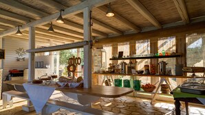 Uma cozinha rústica com vigas de madeira, chão de pedra e diversos utensílios de cozinha e alimentos expostos em prateleiras.