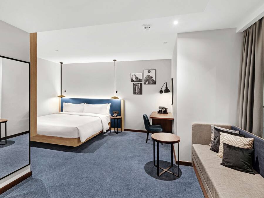 Quarto de hotel moderno com uma cama grande, uma escrivaninha, uma cadeira, uma mesa redonda e um sofá.