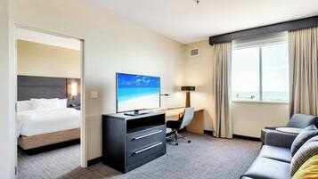 Quarto de hotel com uma cama, uma escrivaninha com um computador, um sofá e uma janela grande com vista para o mar.