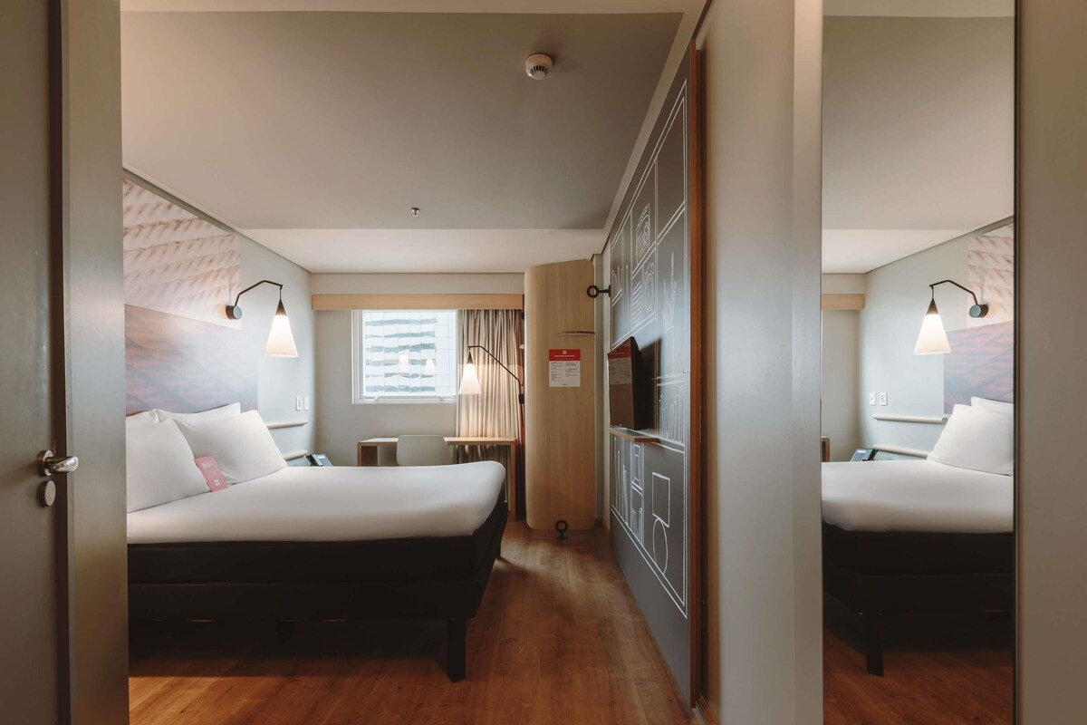 Quarto de hotel com cama, mesa de cabeceira, abajur e espelho.