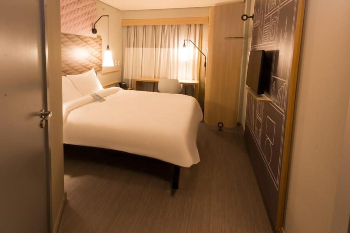 Quarto de hotel com cama, escrivaninha, cadeira, TV e parede com ícones.