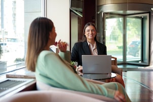 Duas pessoas sentadas em um saguão moderno, com um laptop sobre a mesa, uma divisória de vidro e vista para a rua.