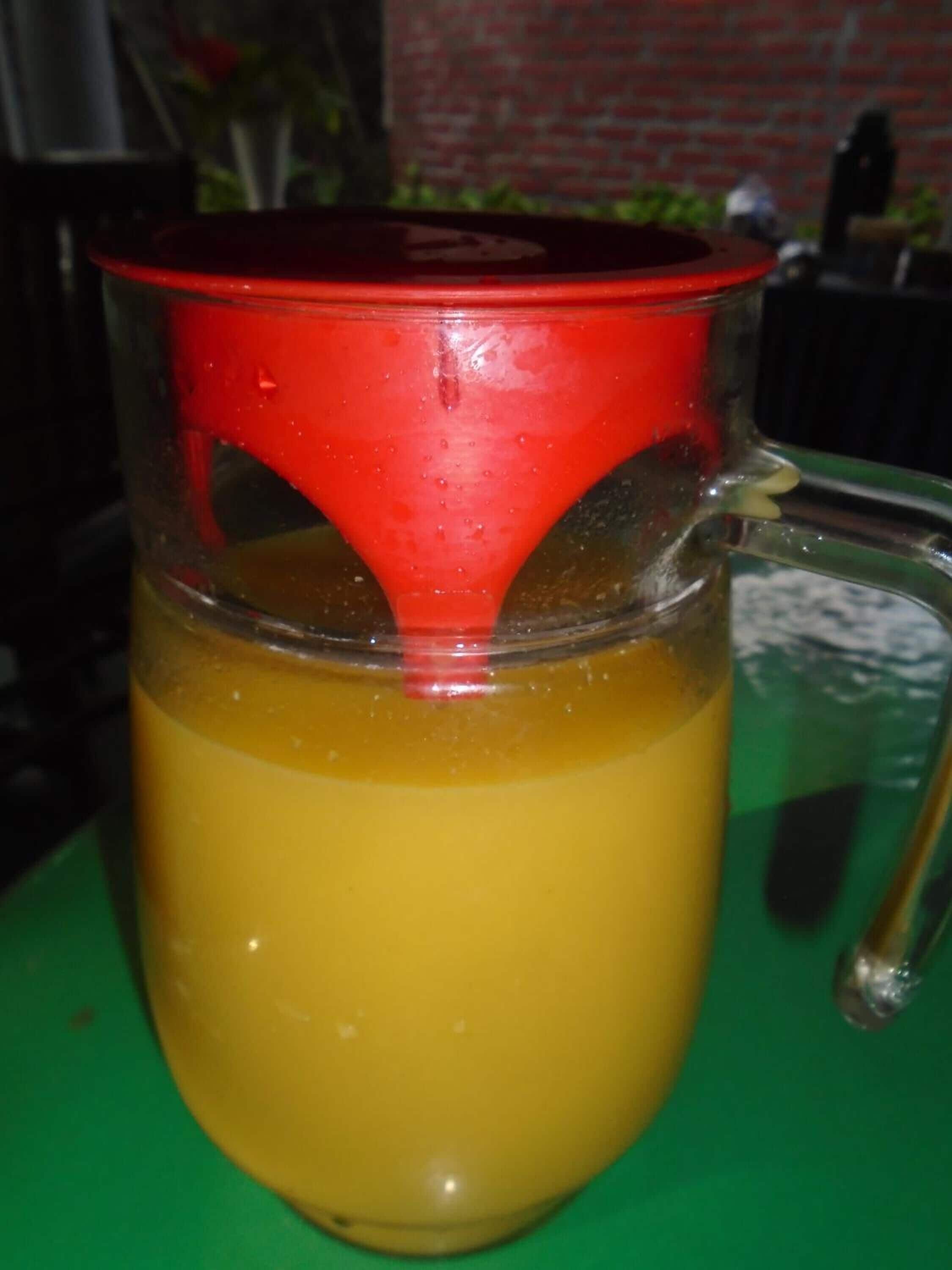 Um jarro de vidro com tampa vermelha, cheio de líquido amarelo, sobre uma superfície verde.