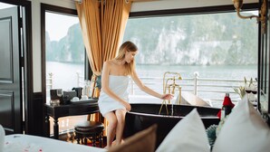 Mulher vestindo um roupão branco toca piano em um quarto com vista para um lago e montanhas.