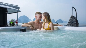Um casal em uma banheira de hidromassagem com vista para o mar e montanhas ao fundo.