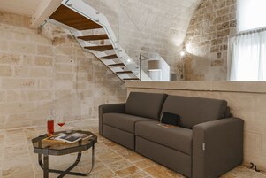 Uma sala de estar moderna com parede de pedra, uma mesa de vidro com uma garrafa e copos, e um sofá cinzento.