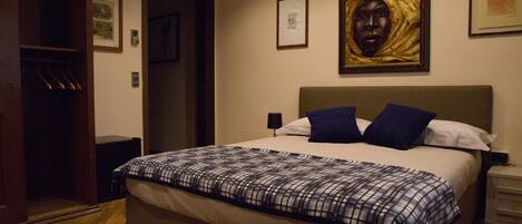 Quarto com cama, mesa de cabeceira, luminária, guarda-roupa de madeira e quadros nas paredes.