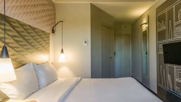 Quarto de hotel com cama, luminária, telefone e televisão fixada na parede.