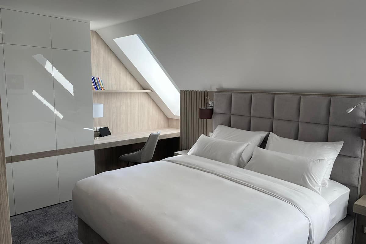 Quarto de hotel moderno com uma cama grande, uma mesa de trabalho e um claraboia.