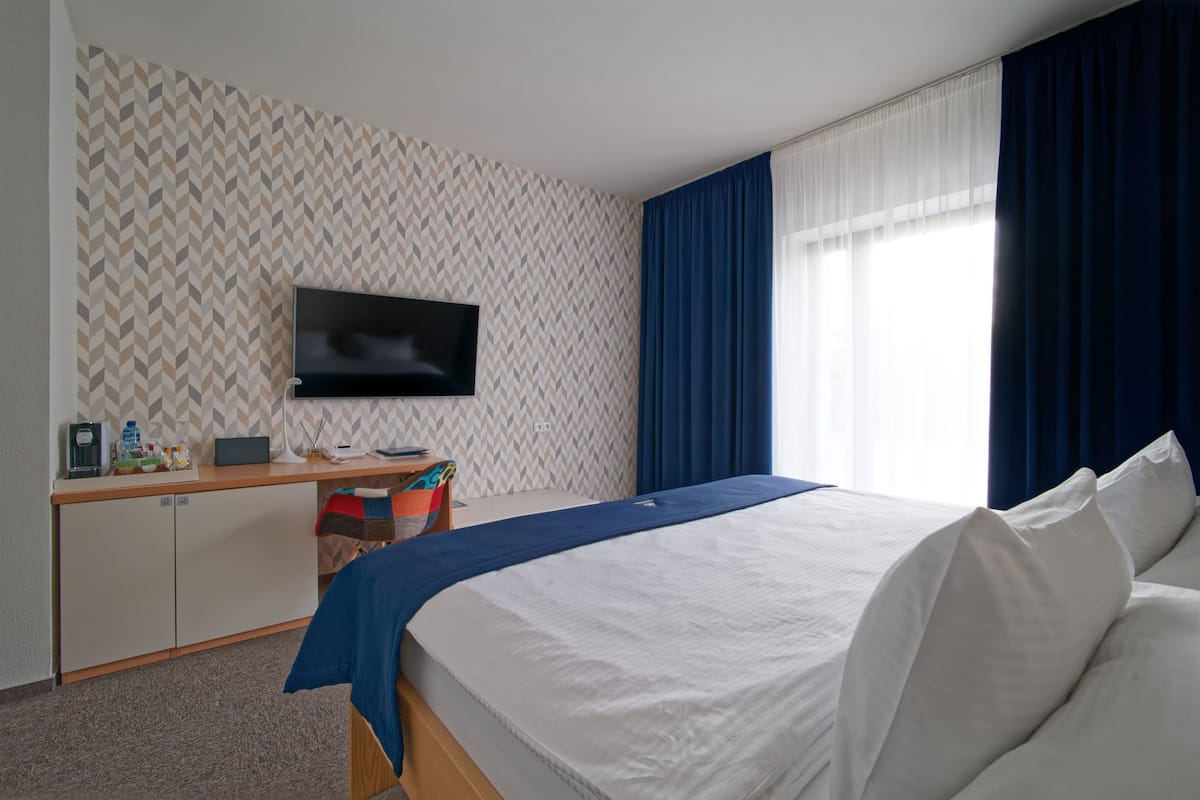 Quarto de hotel com uma cama, uma escrivaninha com televisão, um guarda-roupa e uma janela com cortinas.