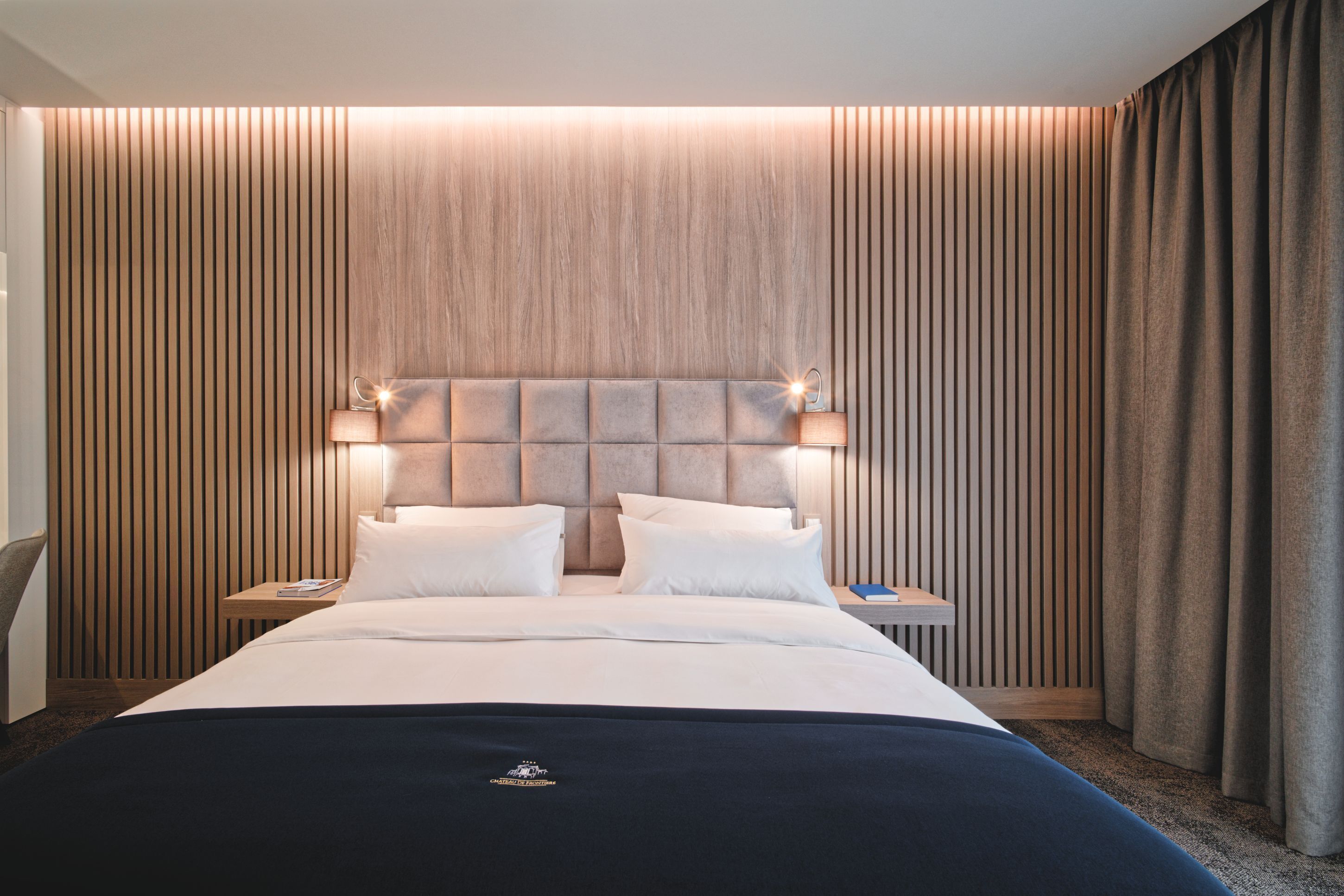 Quarto de hotel com uma cama grande, duas mesas de cabeceira, cabeceira com iluminação integrada e uma cortina no lado direito.