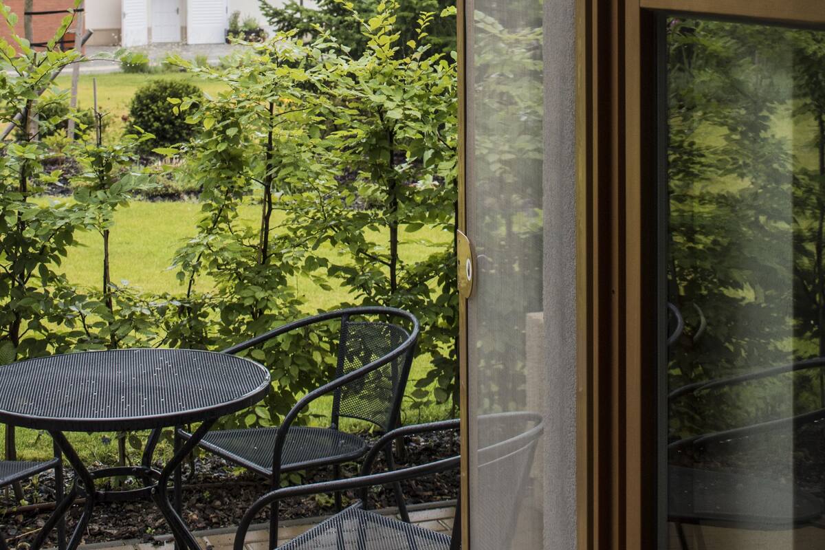 Conservatório com porta de vidro, mesa e cadeiras no pátio, com vista para um jardim e um edifício com fachada branca e janelas em arco.