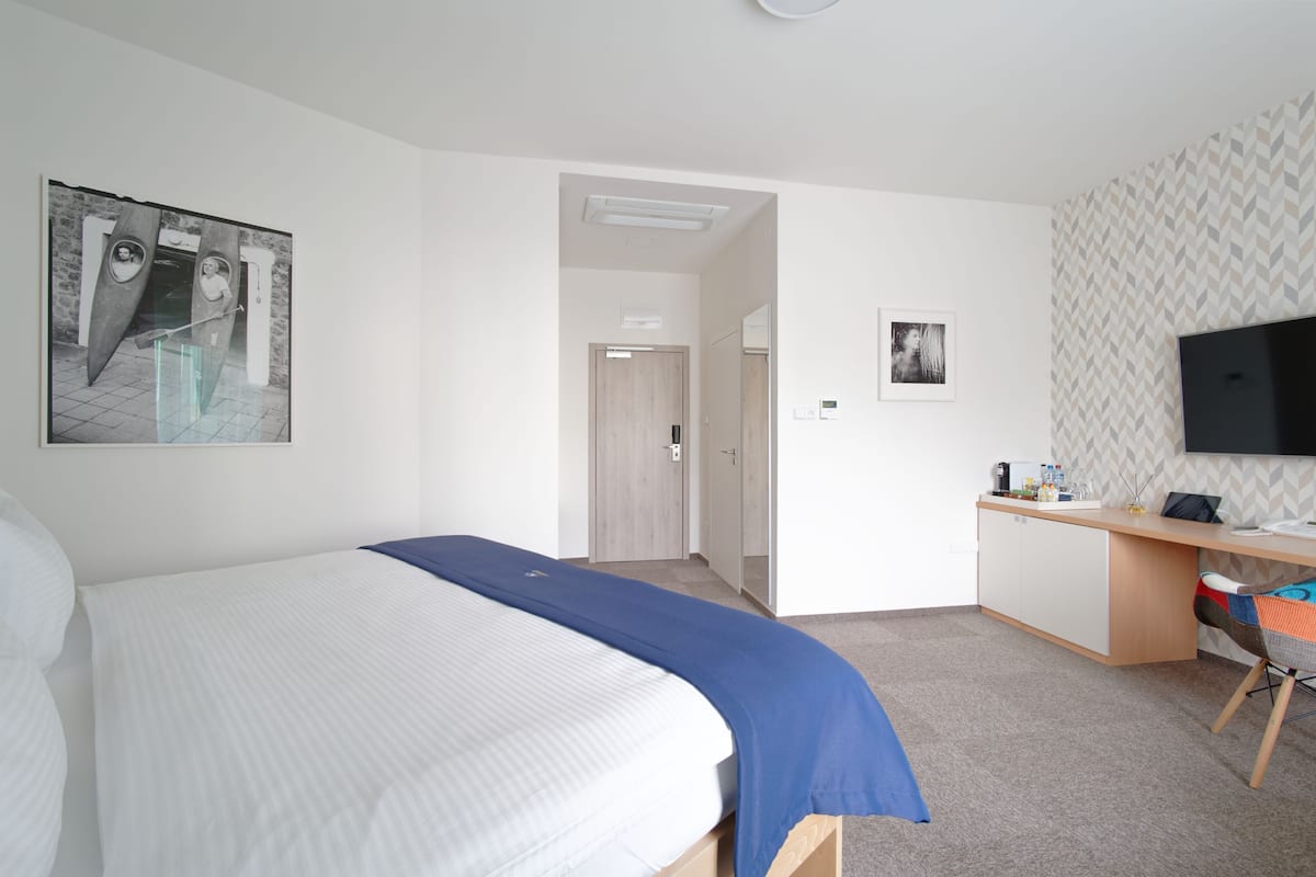 Quarto de hotel com cama, escrivaninha, cadeira, televisão e porta que dá para outro cômodo.