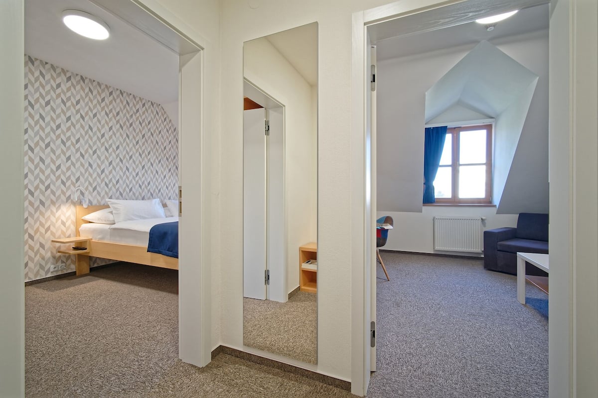 Quarto com cama, mesa de cabeceira e espelho. Há um corredor que leva a outro cômodo com janela e sofá.