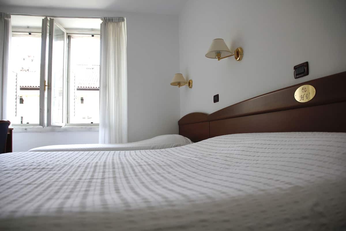Quarto de hotel com duas camas, uma janela com cortinas, luminárias na parede e um relógio na cabeceira.