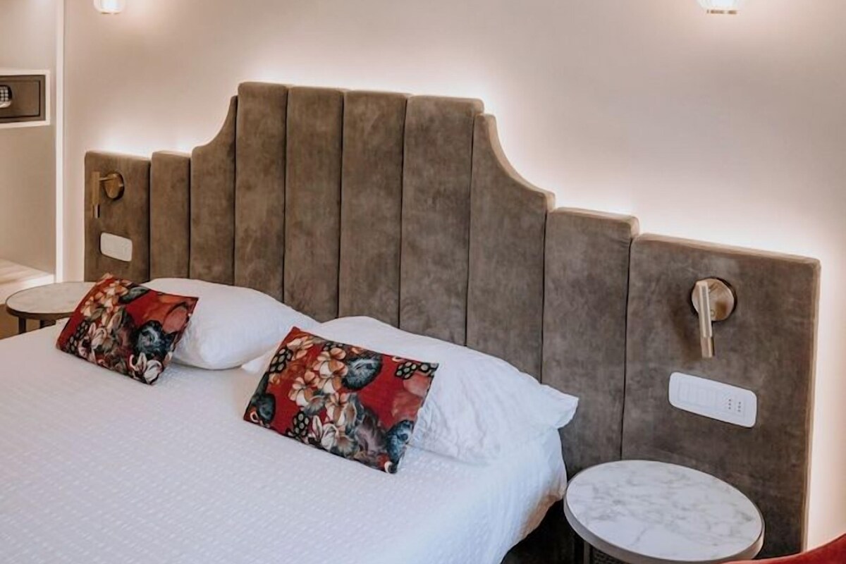 Quarto de hotel com uma cama grande, duas mesas de cabeceira, uma cadeira vermelha e luminárias fixadas na parede.