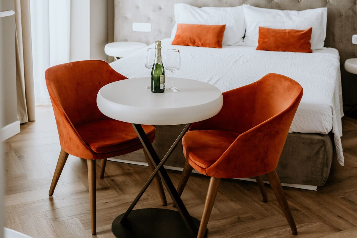 Quarto de hotel moderno com cabeceira estofada, mesa de jantar redonda, duas cadeiras laranja, uma garrafa de champanhe e cama com roupa de cama branca e laranja.