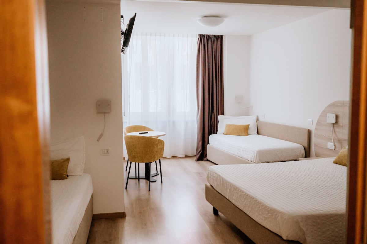Quarto de hotel com duas camas de solteiro, uma mesa pequena, uma cadeira e janela com cortinas.