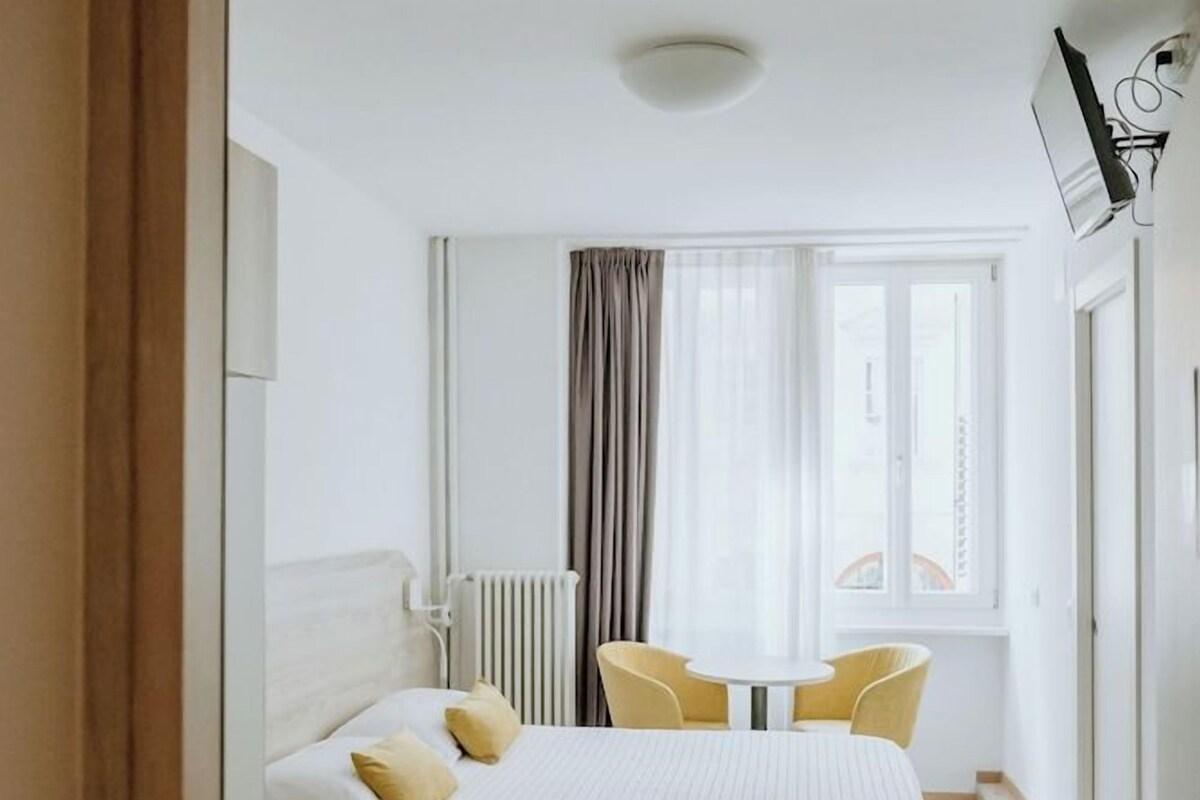 Quarto com uma cama, duas cadeiras, um radiador e uma janela com cortinas.