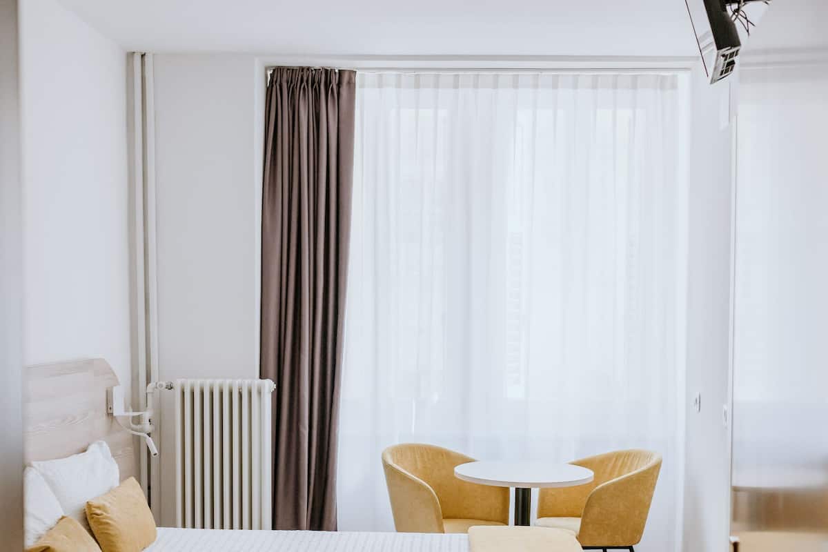Quarto de hotel com cama, uma pequena mesa com cadeiras, radiador e janela com cortinas.