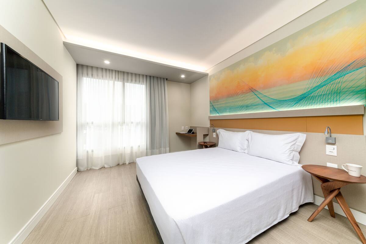 Quarto de hotel com uma cama grande, televisão de tela plana na parede, mesa de cabeceira com uma xícara e um quadro acima da cama.